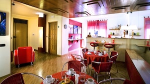 Uma área de refeições de hotel com cadeiras e mesas vermelhas, piso de madeira e uma cozinha ao fundo.