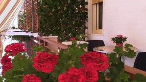 Uma varanda com corrimão, uma mesa e cadeiras. Há plantas e flores em vasos sobre a mesa.