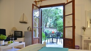 Quarto com cama, televisão, uma mesinha com uma planta, e vista para um pátio com cadeiras e mesa.