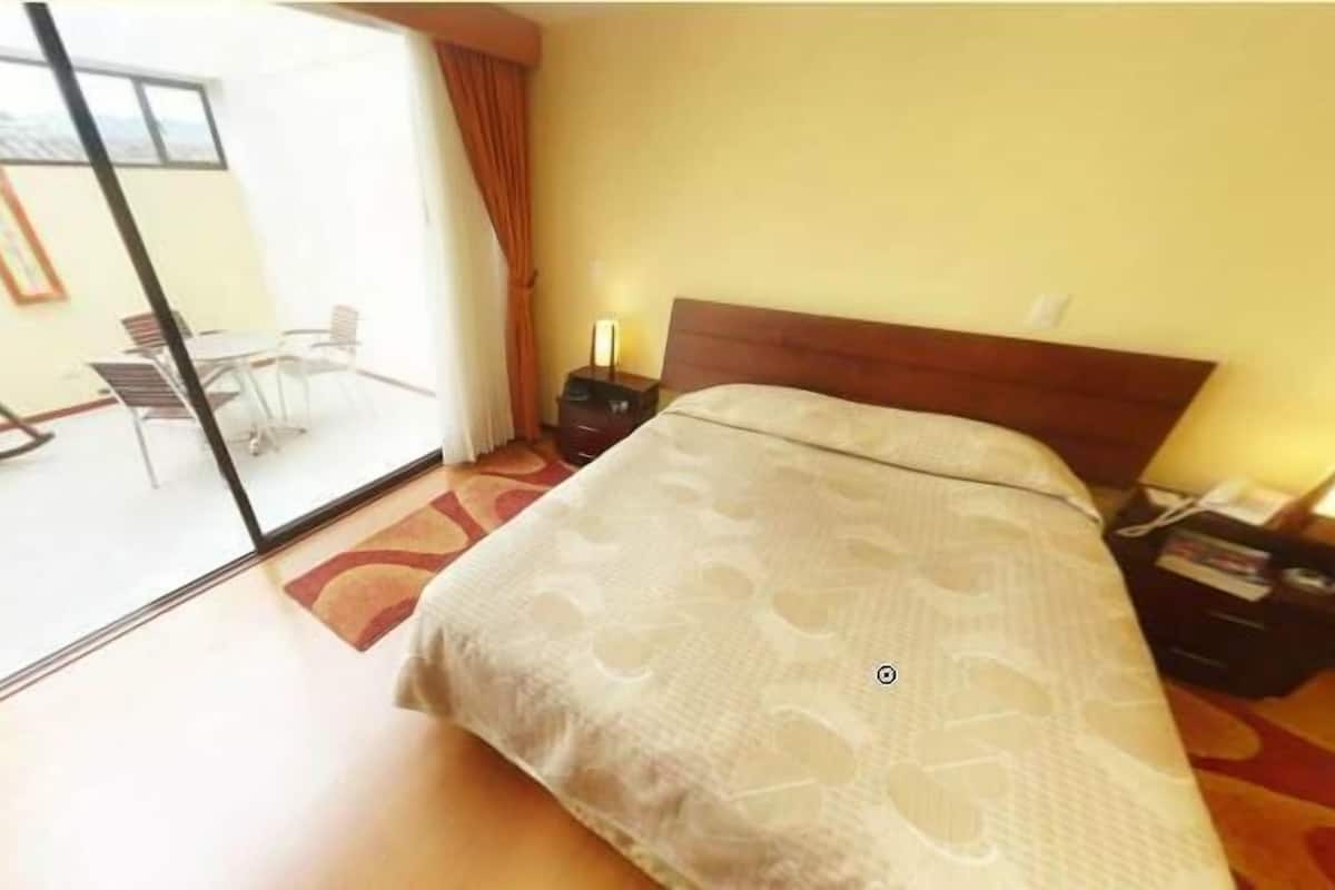 Quarto de hotel com cama, mesa de cabeceira, escrivaninha com cadeiras e vista para área externa com cadeiras e mesa através de uma porta deslizante.