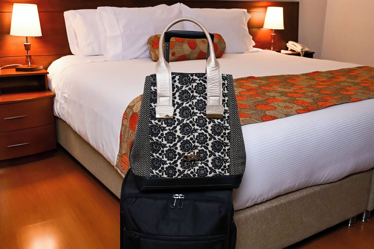 Quarto de hotel com uma cama, mesa de cabeceira, abajur, mala e bolsa.