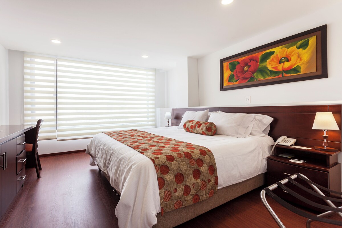 Quarto de hotel com cama, mesa de cabeceira, luminária, cadeira, escrivaninha e janela com persianas.