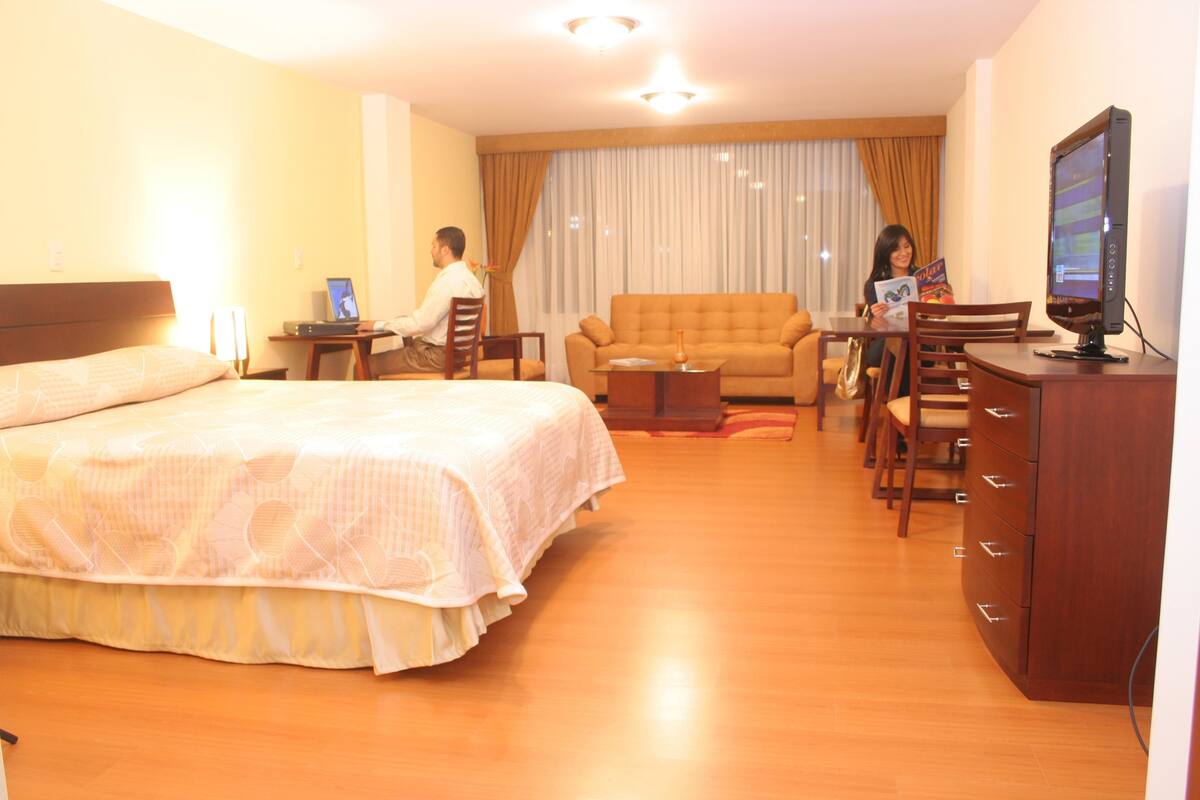 Quarto de hotel com uma cama grande, um sofá, uma televisão e uma mesa de escritório com um laptop.