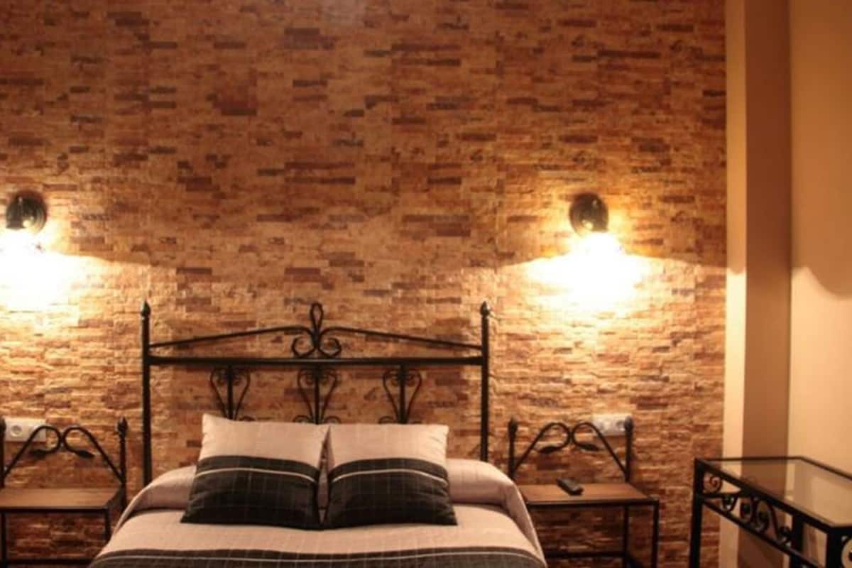 Quarto com parede de tijolos, estrutura de cama de metal, duas mesas de cabeceira e duas luminárias fixadas na parede.