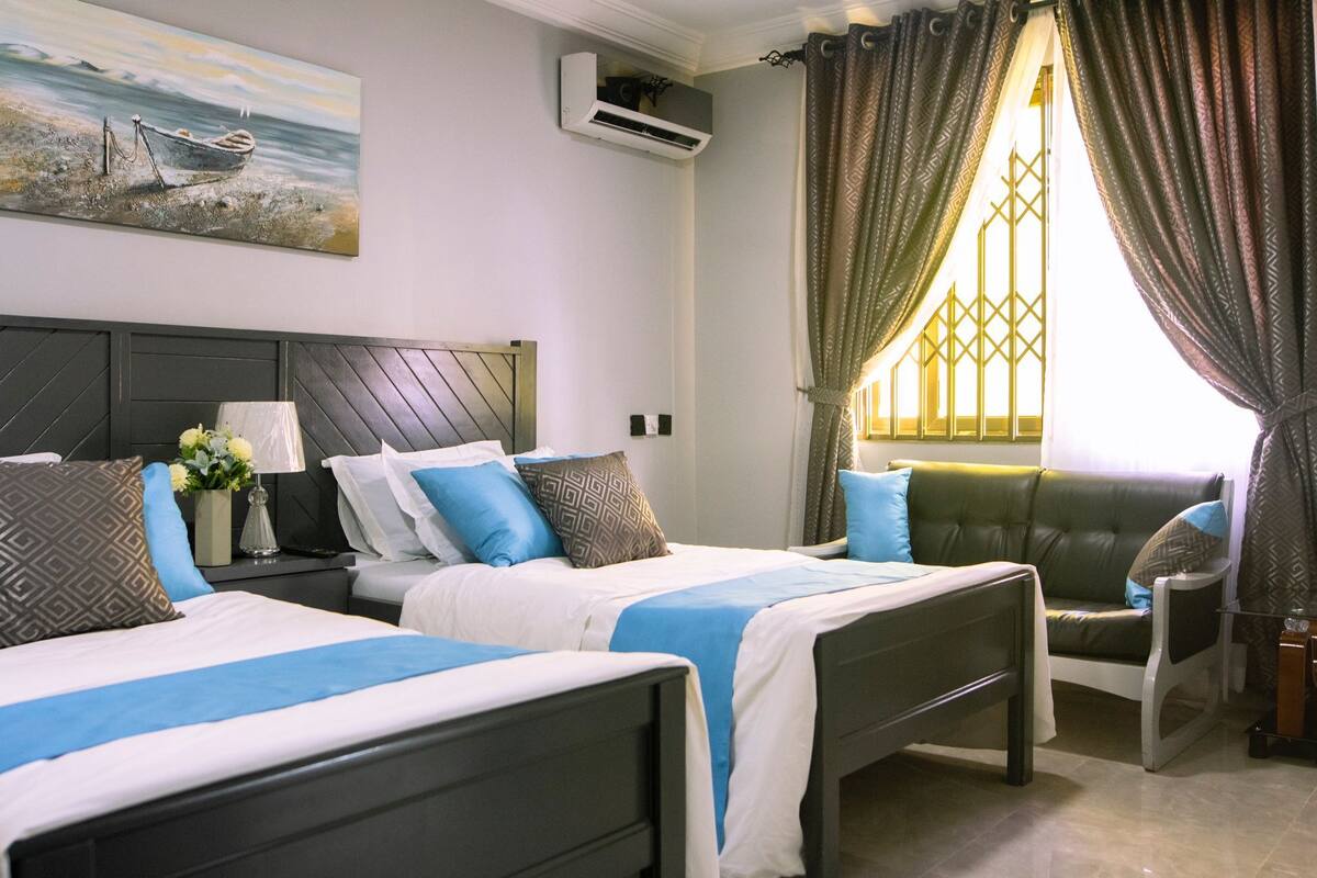 Quarto de hotel com duas camas, um sofá verde, uma janela decorativa com cortinas estampadas e um ar condicionado fixado na parede.