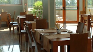 Um restaurante com mesas e cadeiras de madeira, janelas amplas e vista para o verde lá fora.