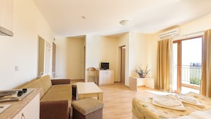 Quarto de hotel com sofá, uma mesa pequena, televisão, varanda com cortinas e ar condicionado.