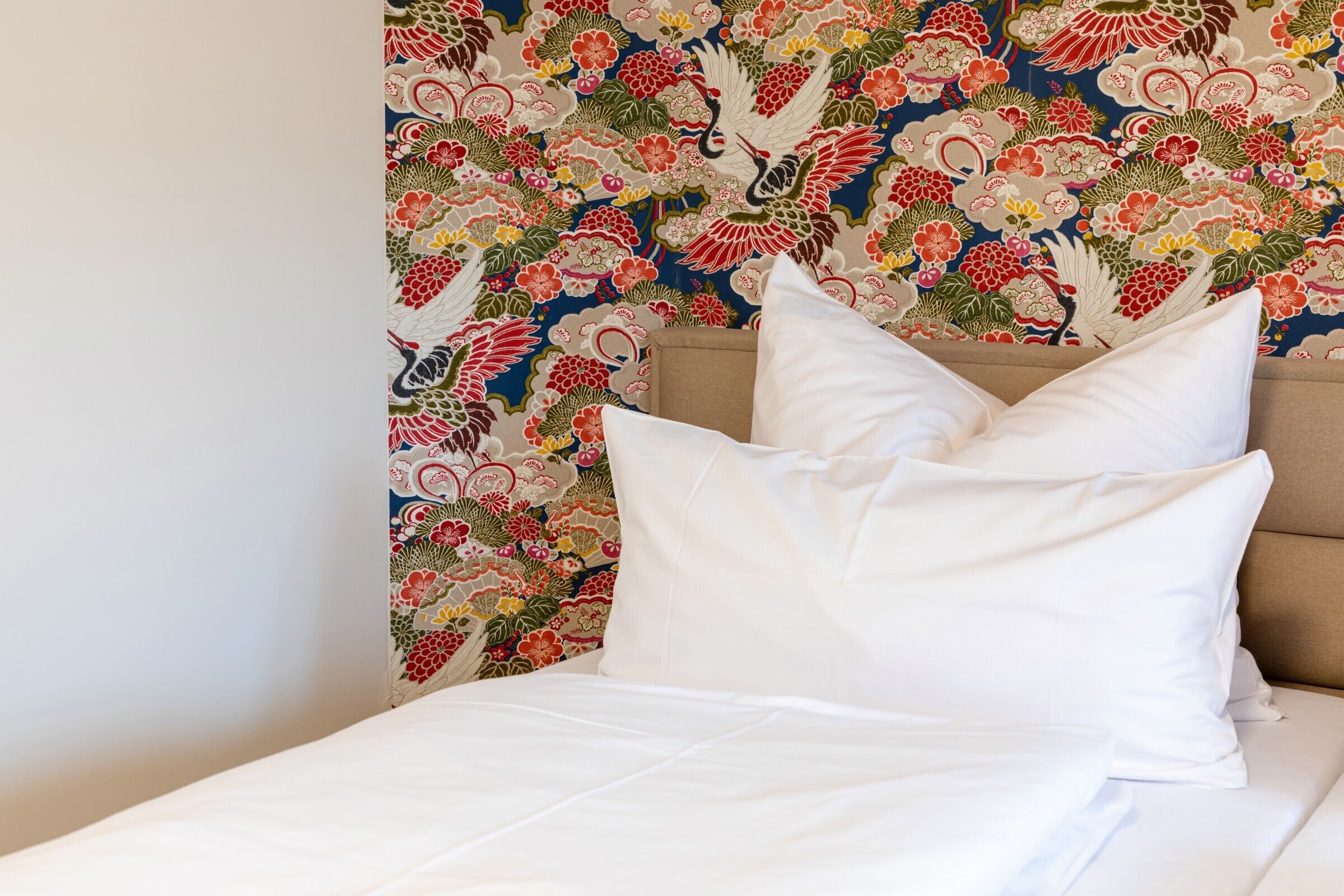 Cama com roupa de cama branca e duas almofadas, diante de uma parede com papel de parede estampado com flores.