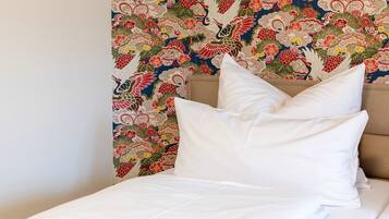 Cama com roupa de cama branca e duas almofadas, diante de uma parede com papel de parede estampado com flores.