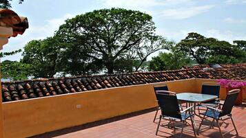 Um terraço com telhado em azulejos, uma mesa com cadeiras e vista para árvores e um corpo de água.