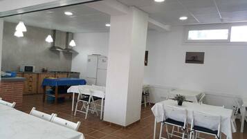 Uma área de refeições com mesas e cadeiras, uma cozinha ao fundo e uma janela.