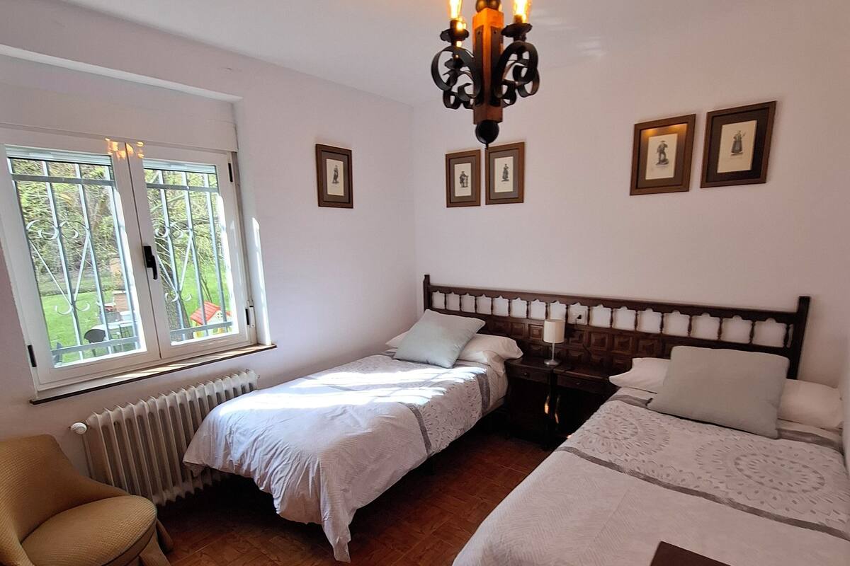 Quarto com duas camas de solteiro, piso de madeira, radiador, cadeira e janela com vista para área verde.