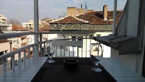 Varanda com mesa posta para dois, com vista para área residencial com edifícios de tijolo e telhados de telha vermelha.