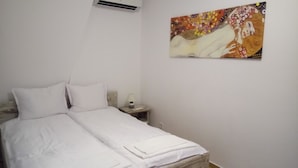 Uma cama de solteiro com roupa de cama branca num quarto com parede branca lisa. Há uma pequena mesa de cabeceira com uma lâmpada e uma tomada. Um quadro colorido está pendurado na parede acima da cama.