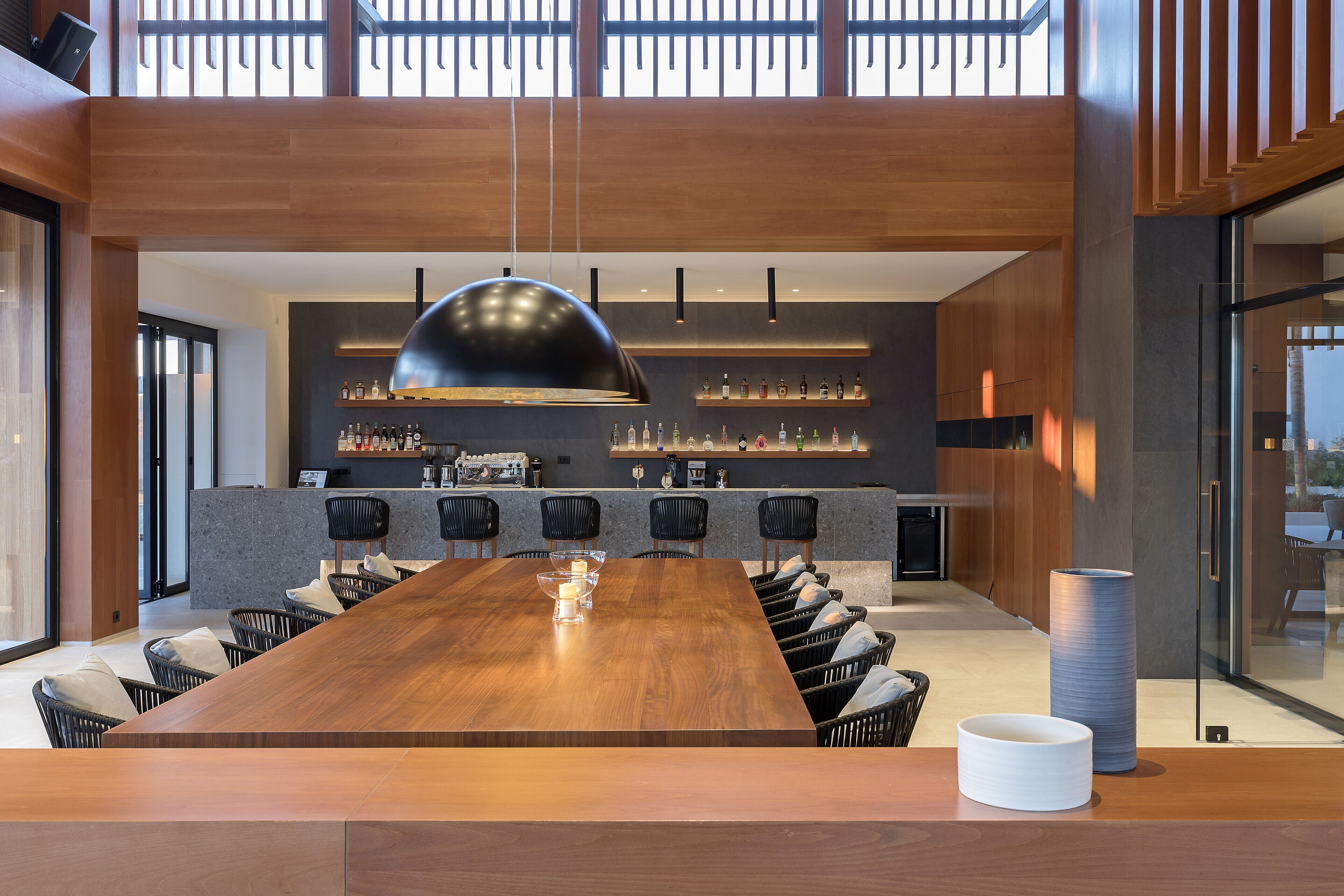 Uma sala de jantar moderna com uma mesa de madeira comprida, cadeiras pretas e uma luminária pendente central.