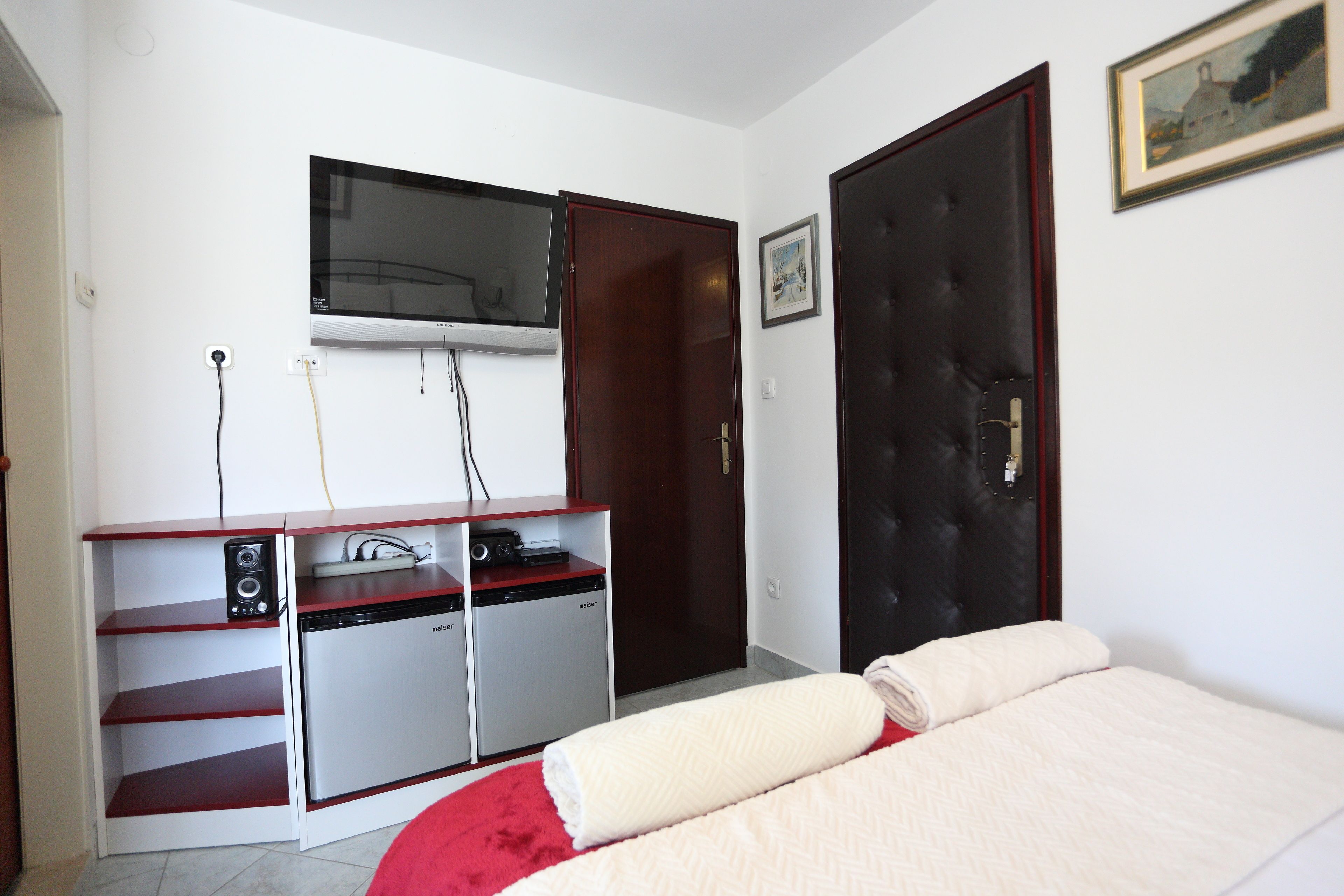 Um quarto com cama, guarda-roupa, escrivaninha e televisão, além de uma porta.