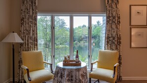 Quarto com duas poltronas amarelas, uma mesa com toalha de mesa florida, um abajur e vista para um lago através de grandes janelas.