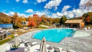 Piscina com espreguiçadeiras e guarda-sóis, cercada por árvores de outono e vista para um lago.