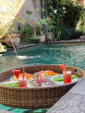 Uma bandeja de vime com bebida, uma vela e petiscos junto a uma piscina.