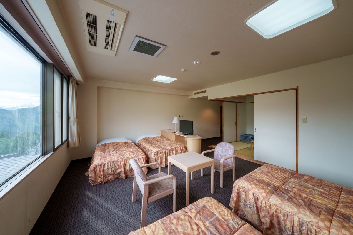 Quarto de hotel com duas camas, uma mesa, uma cadeira, televisão e janela com vista para as montanhas.