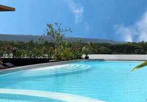 Piscina com água azul cristalina, cercada por plantas tropicais e vista para montanhas ao longe.