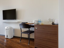 Quarto de hotel com secretária, cadeira, televisão de tela plana, cafeteira, garrafas de água e lixeira.