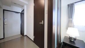 Quarto de hotel com porta fechada, mesa de cabeceira com abajur e janela com cortinas.