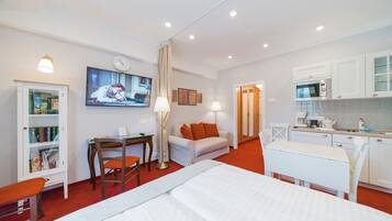 Quarto de hotel com cama, escrivaninha, cadeira, estante de livros, televisão, sofá e cozinha compacta.