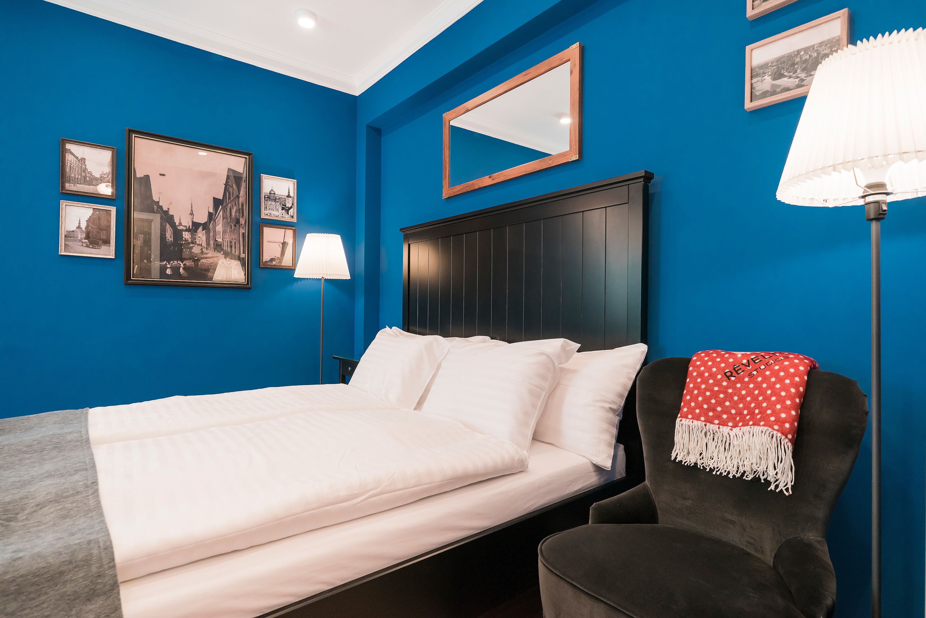 Quarto de hotel com cama, uma cadeira, abajur e quadros na parede.