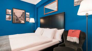 Quarto de hotel com cama, uma cadeira, abajur e quadros na parede.