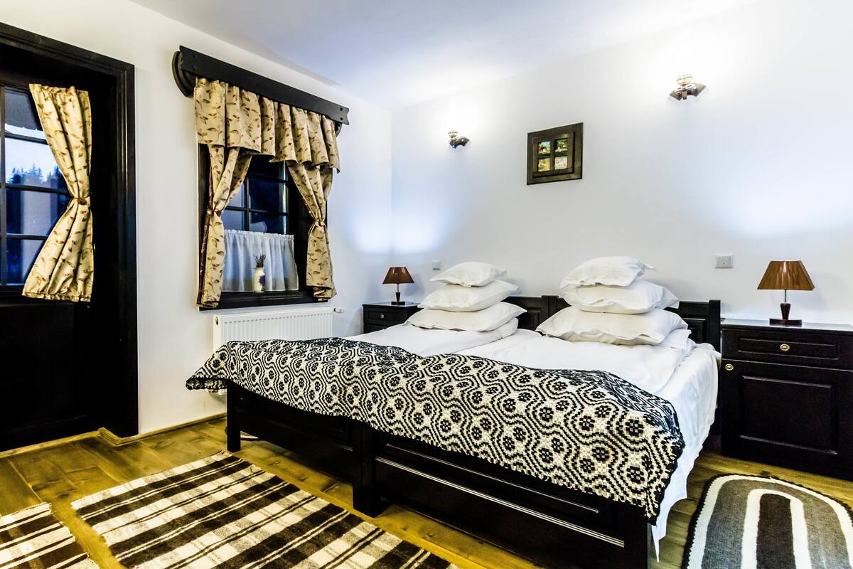 Quarto com cama, duas mesas de cabeceira, janela com cortinas, radiador e um quadro na parede.