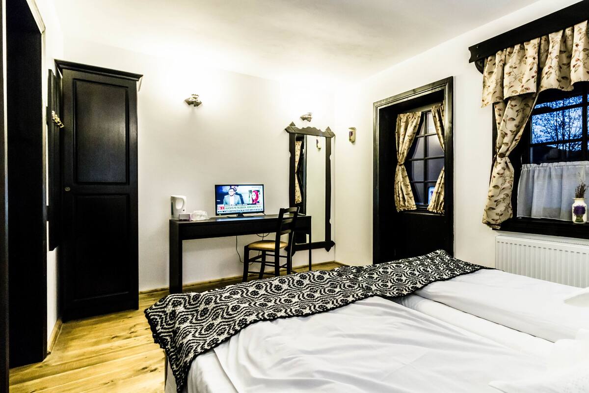Quarto com cama, escrivaninha com computador, cadeira, janela com cortinas e radiador.