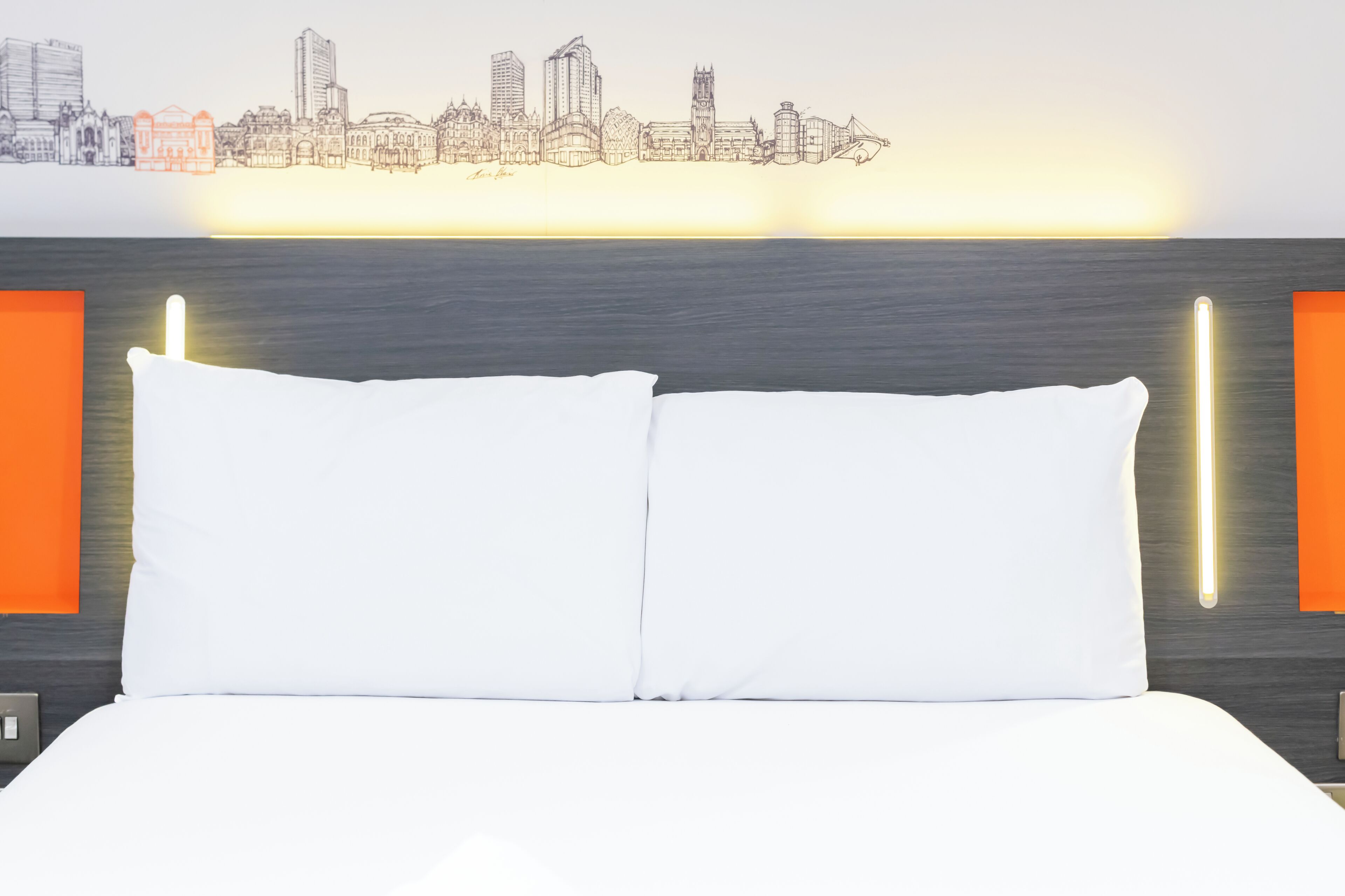 Quarto de hotel com cama, almofadas brancas, mural da paisagem urbana na parede e iluminação em tons de laranja.