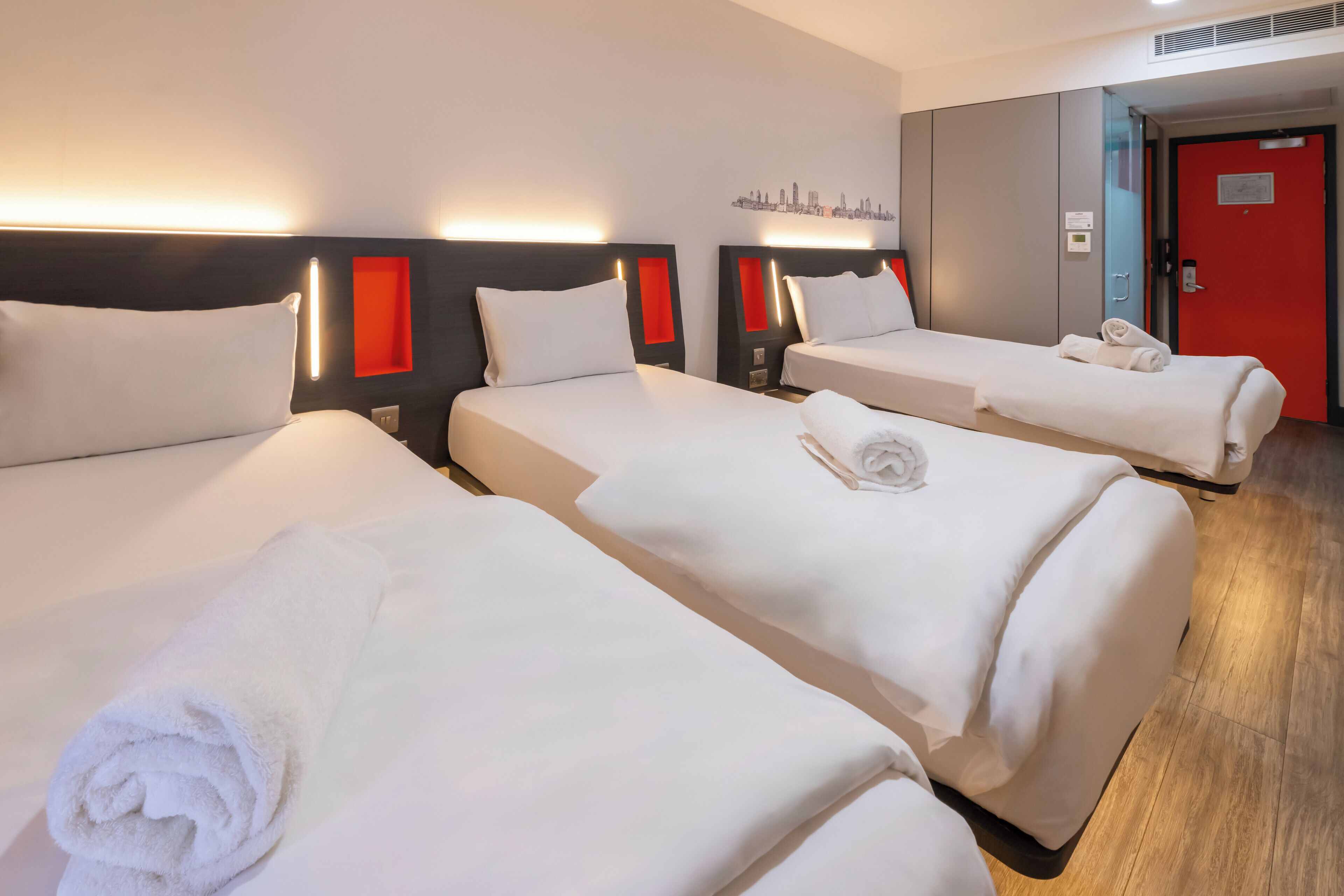 Quarto de hotel com três camas de solteiro, cada uma com roupa de cama branca e toalha dobrada. O quarto possui cabeceiras modernas com iluminação integrada e uma parede de destaque vermelha.