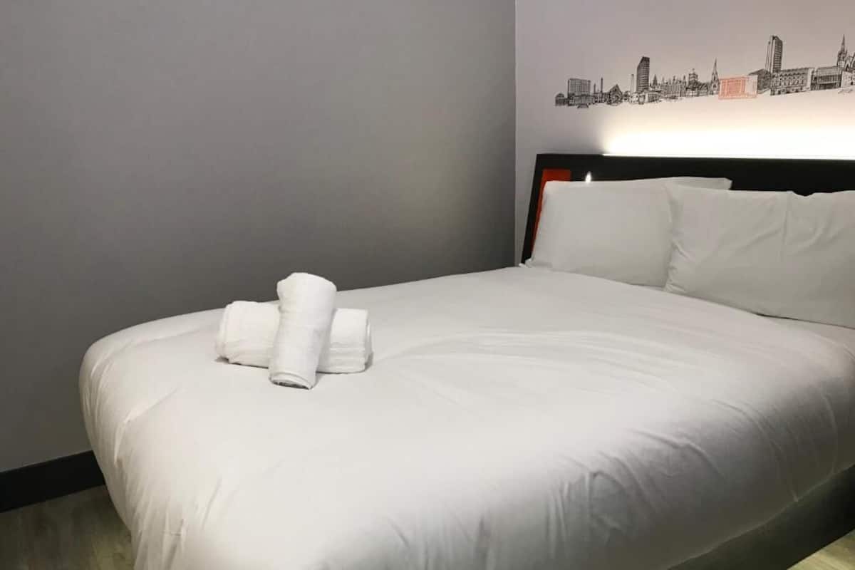 Cama bem arrumada com lençóis brancos e uma pintura de paisagem urbana na parede.