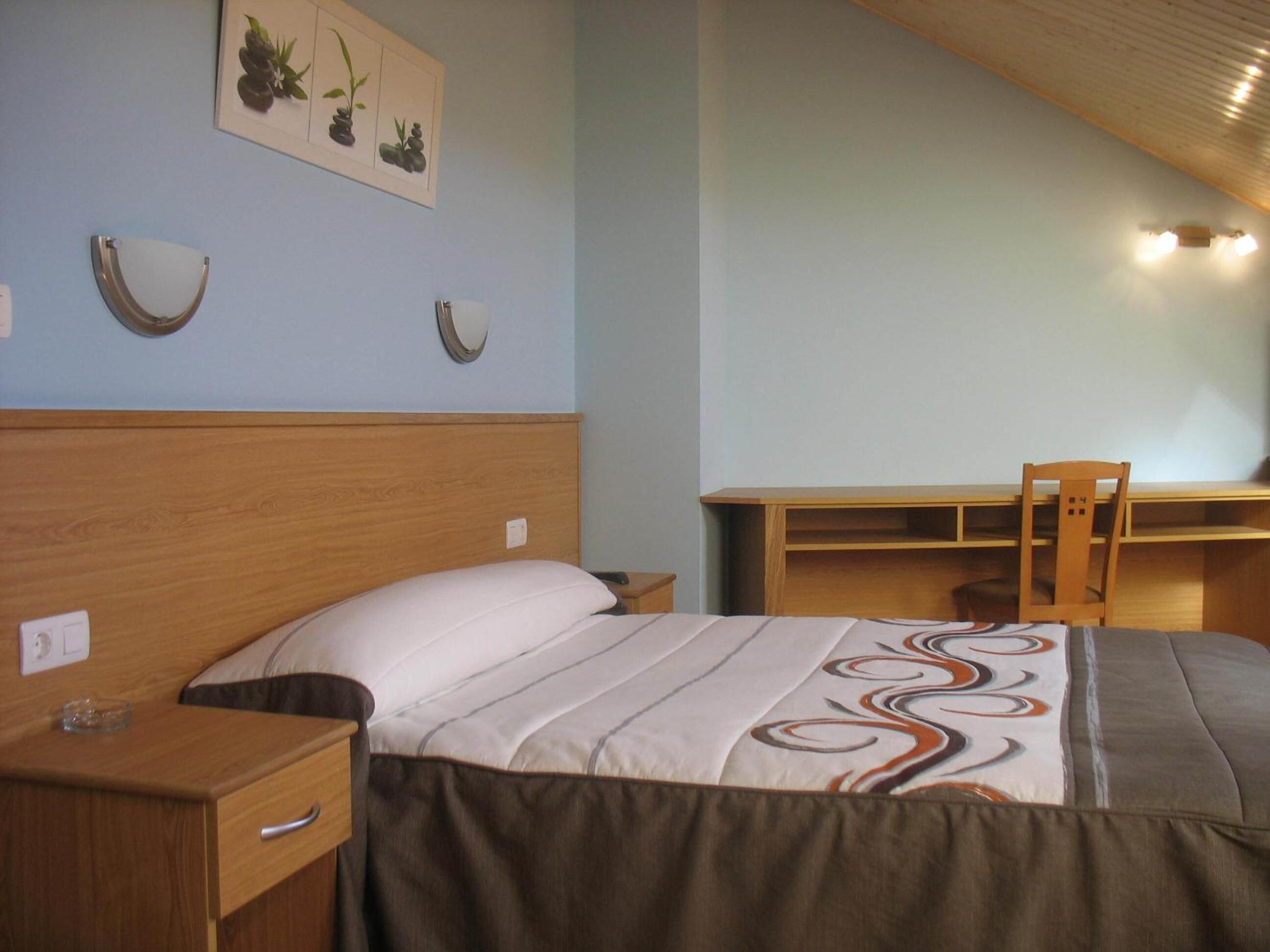 Quarto com cama de madeira, escrivaninha e cadeira. Há uma prateleira na parede e um quadro emoldurado acima da cama.