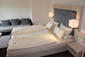 Uma cama de casal com roupa de cama branca, cabeceira com desenho estampado, duas luminárias de cabeceira e uma mesa de cabeceira com abajur.
