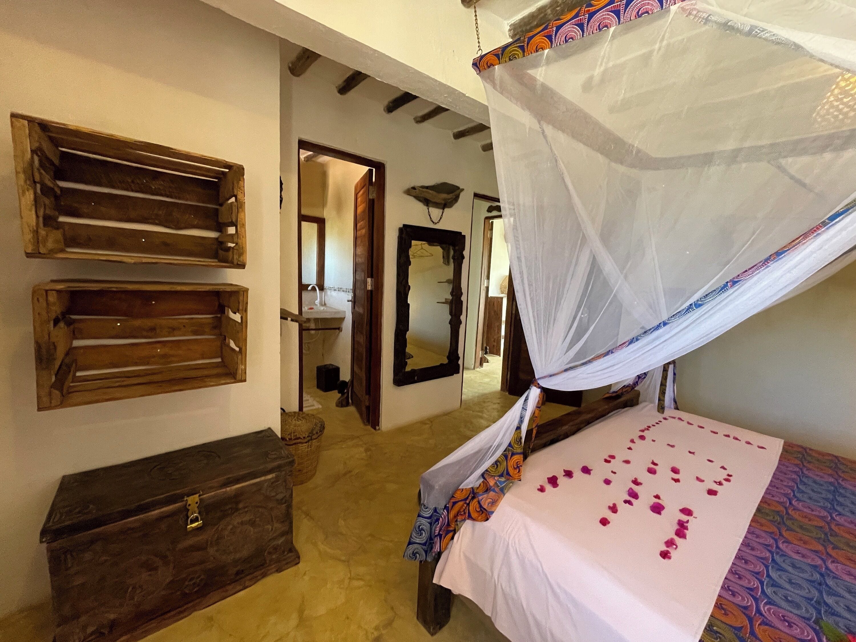 Quarto com cama de dossel, cabeceira de madeira, cômoda, espelho e porta que dá para outro ambiente.