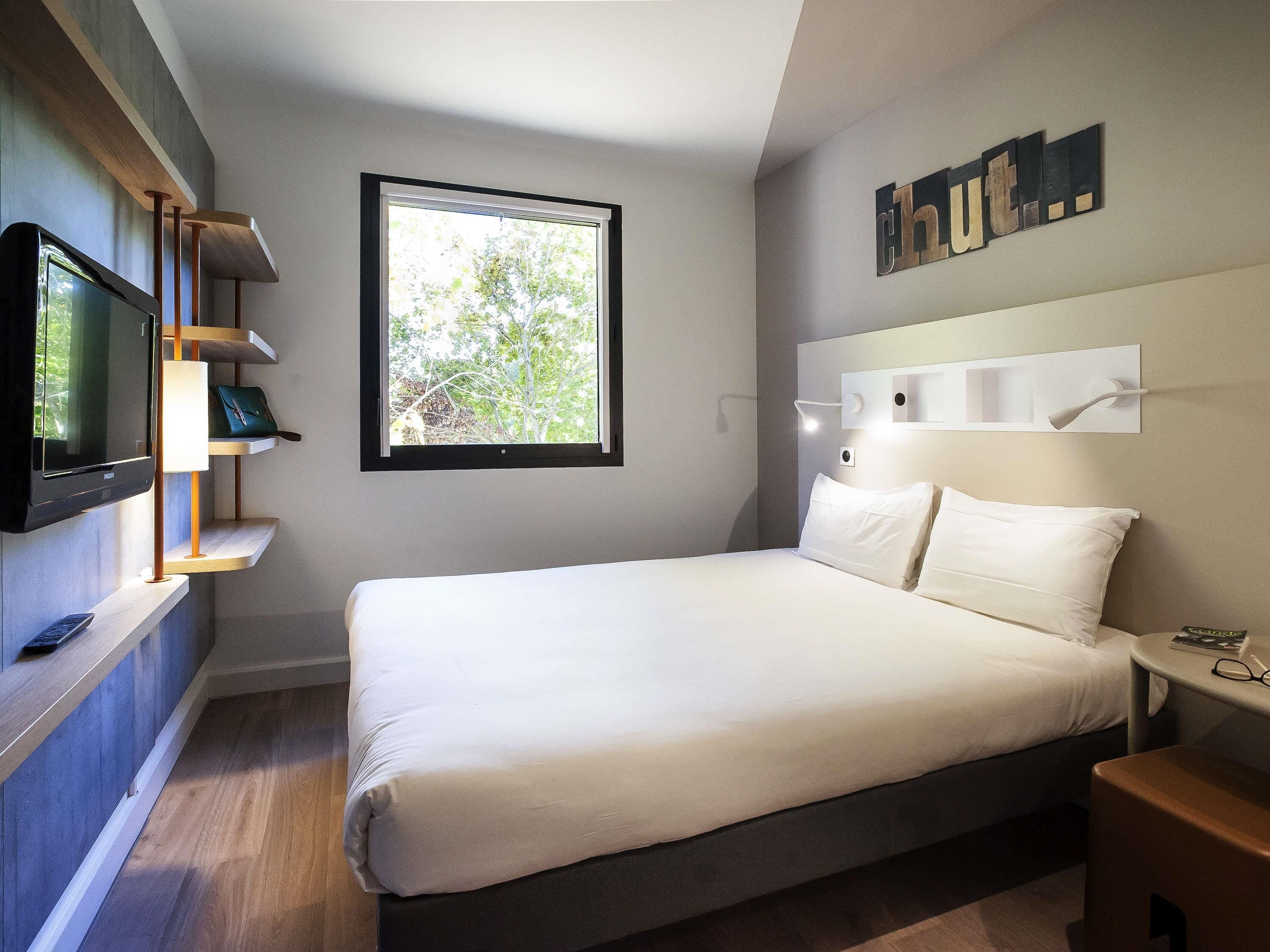 Quarto de hotel moderno com uma cama grande, televisão fixada na parede, janela com vista para árvores e design minimalista.