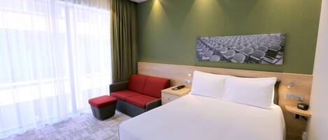Quarto de hotel com uma cama grande, um sofá vermelho, uma mesa de cabeceira e um quadro pendurado na parede.