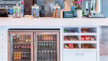Uma área de recepção de hotel com um minibar, geladeira abastecida com bebidas e uma prateleira com petiscos.