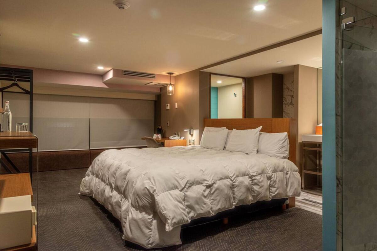 Quarto de hotel moderno com uma cama grande, mesas de cabeceira, um espelho e área de chuveiro.
