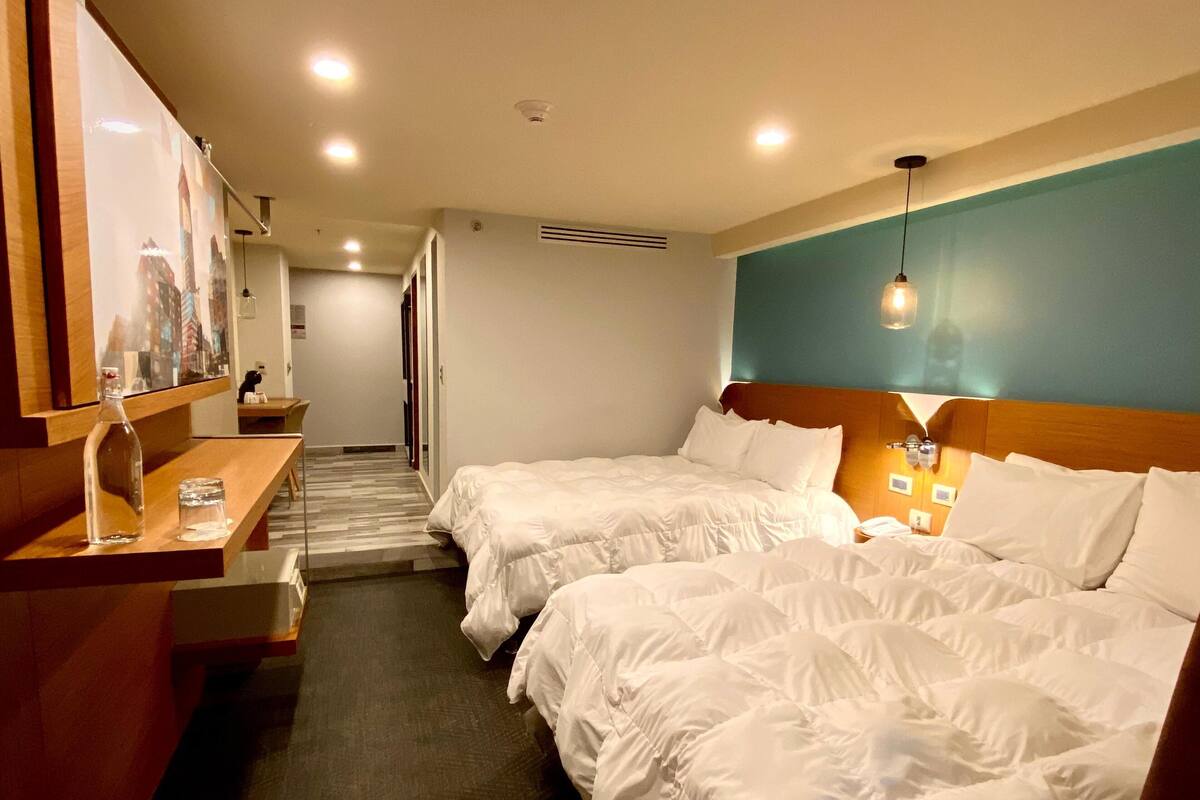 Quarto de hotel com duas camas, uma escrivaninha de madeira, uma mesa de cabeceira, uma luminária na parede e um quadro.