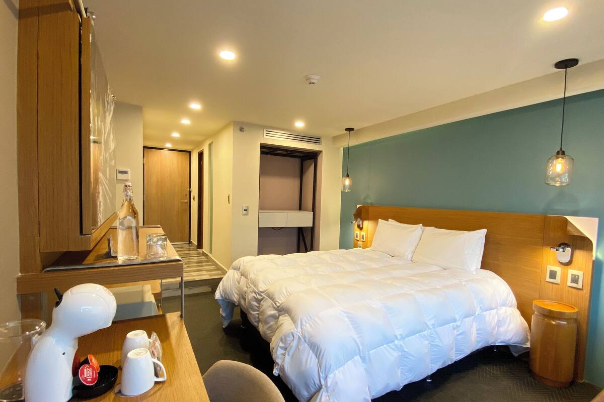 Quarto de hotel com uma cama grande, uma escrivaninha de madeira com cafeteira, um espelho e vista para o corredor.