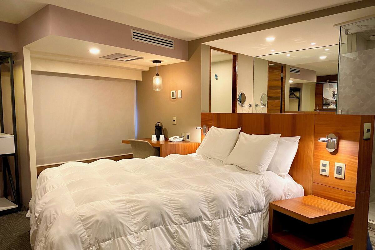 Quarto de hotel com uma cama grande, uma escrivaninha, uma cadeira, um espelho e uma pequena mesa de cabeceira de madeira.