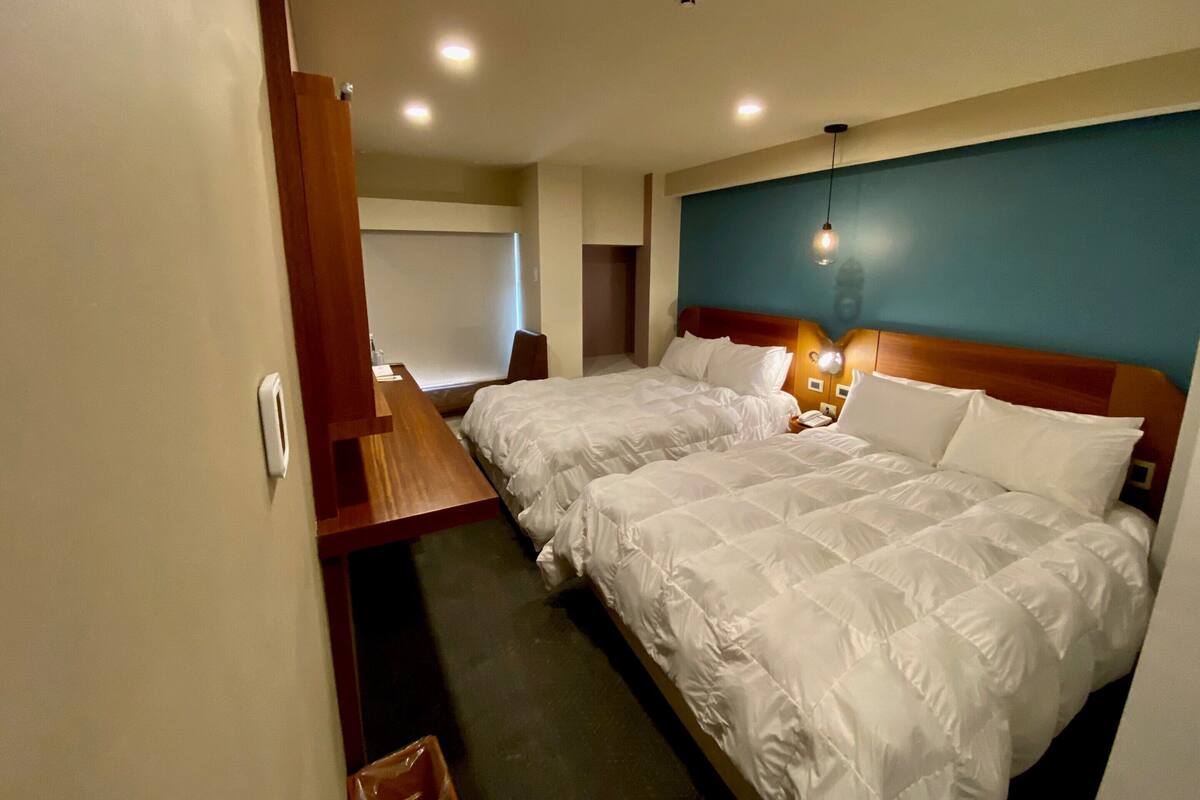 Quarto de hotel com duas camas, uma escrivaninha e uma janela.