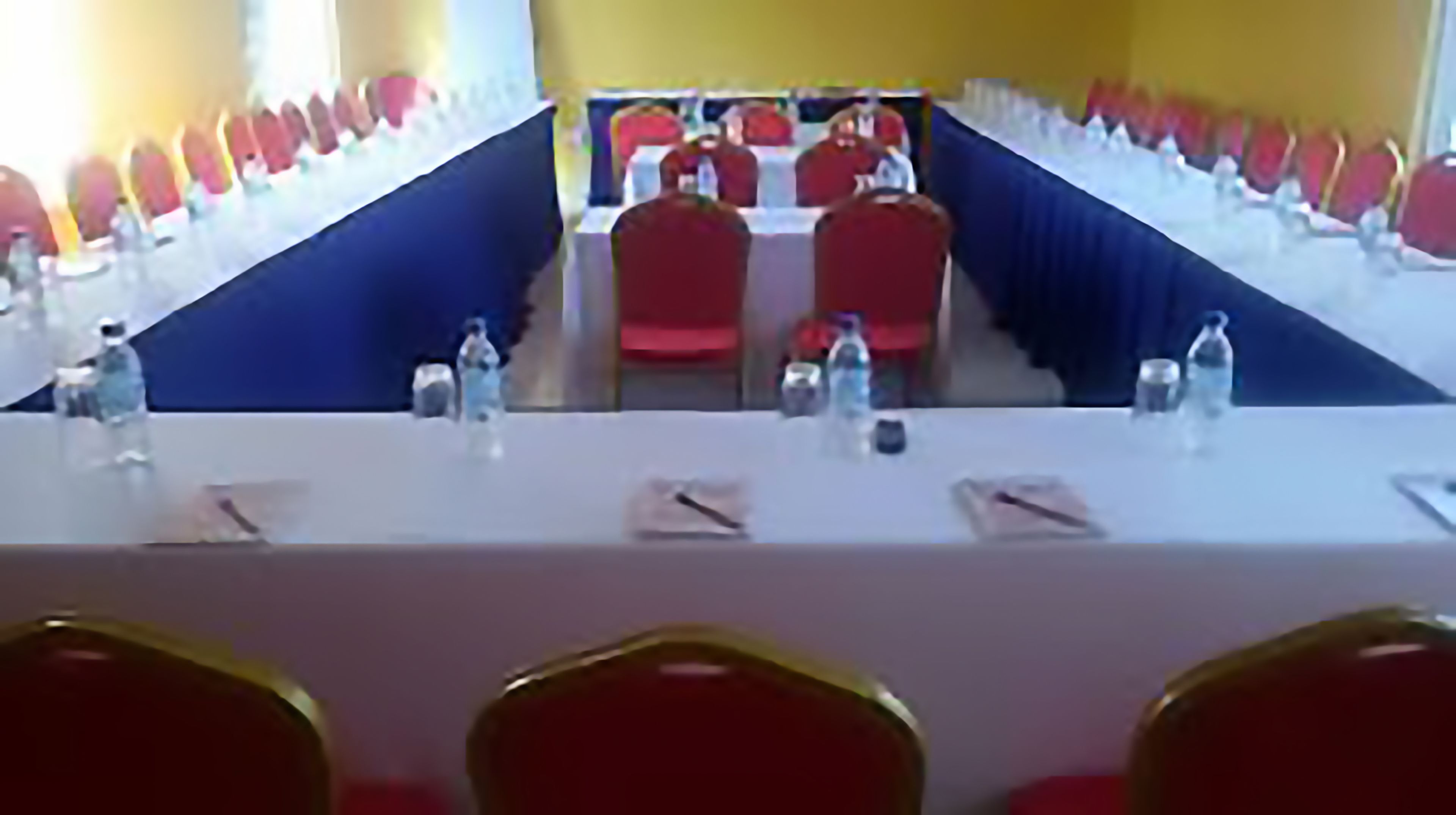 Sala de conferências com uma mesa comprida, cadeiras vermelhas e garrafas de água.