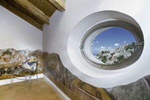 Quarto com janela redonda que oferece vista para uma vila no sopé da colina.