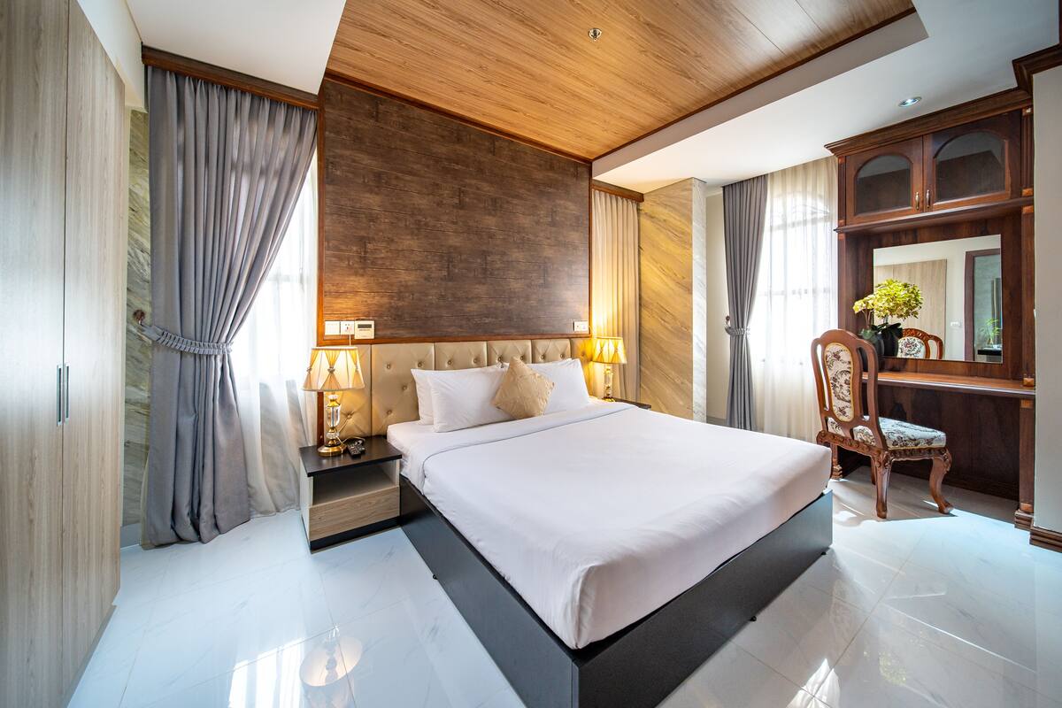 Quarto de hotel moderno com uma cama grande, cabeceira de madeira e uma mesa de cabeceira com abajur.