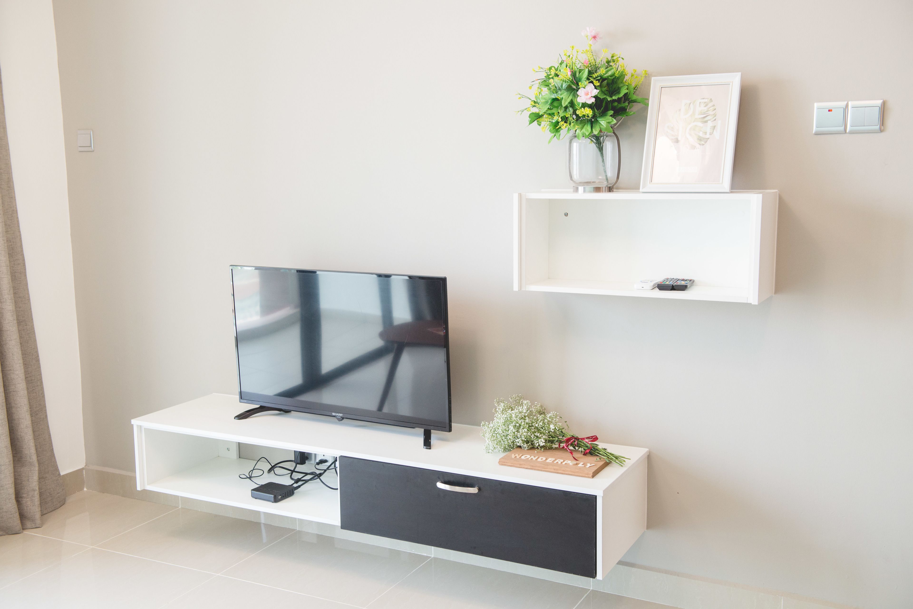 Uma sala de estar moderna com televisão de tela plana sobre um móvel branco, uma pequena planta e um quadro em uma prateleira fixada na parede.